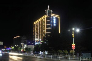 Hotel de vários andares com letreiros iluminados, uma estrutura vertical imponente e vista da rua com cerca e postes de luz.