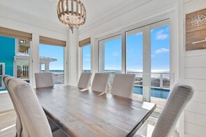 Uma sala de jantar com mesa e cadeiras de madeira, janelas amplas com vista para o oceano e um lustre decorativo.