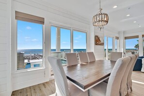 Uma sala de jantar com uma grande mesa de madeira e cadeiras brancas, janelas amplas com vista para o mar e um lustre.