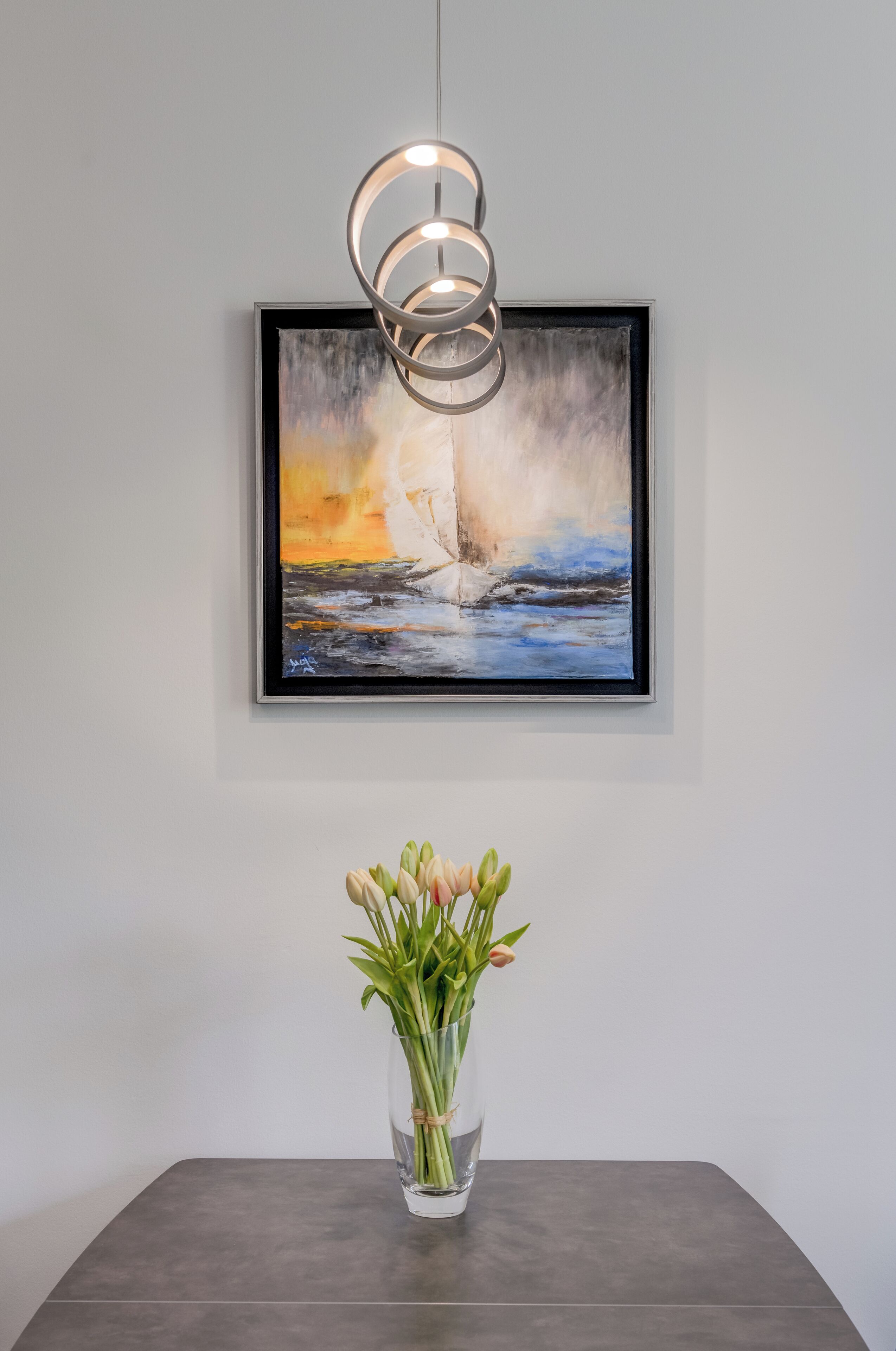 Quadro com um veleiro ao pôr do sol, vaso de vidro transparente com flores sobre uma mesa e uma luminária pendente moderna.