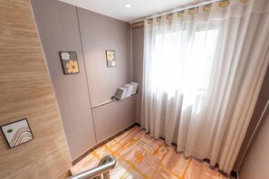Quarto de hotel com piso de madeira, uma cama, janela com cortinas e uma prateleira na parede com alguns objetos.