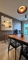 Cozinha moderna com mesa de jantar de madeira, cadeiras pretas, luminárias pendentes e uma televisão.