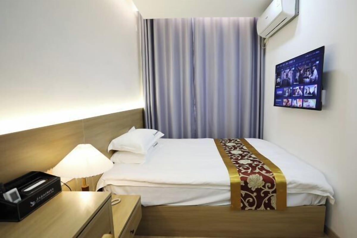 Quarto de hotel com cama, luminária de cabeceira, televisão fixada na parede e janela com cortinas.