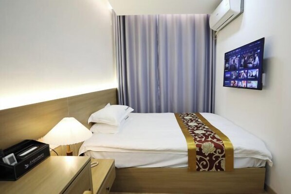 Quarto de hotel com cama, luminária de cabeceira, televisão fixada na parede e janela com cortinas.