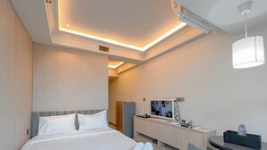 Quarto de hotel moderno com uma cama bem arrumada, uma televisão, uma escrivaninha com uma cadeira e uma pequena mesa com uma garrafa de água.