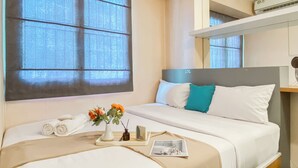 Quarto de hotel com uma janela grande, cama com roupa de cama branca e almofadas verde-água, mesa de cabeceira com um vaso de flores, um quadro e um livro pequeno.