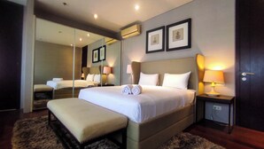 Quarto de hotel com uma cama grande, um banquinho, uma mesinha de cabeceira com luminária e um espelho na parede.