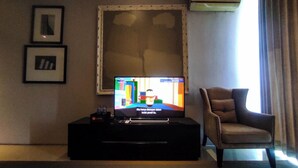 Televisão em suporte preto exibindo um jogo, uma poltrona marrom e duas quadros na parede.