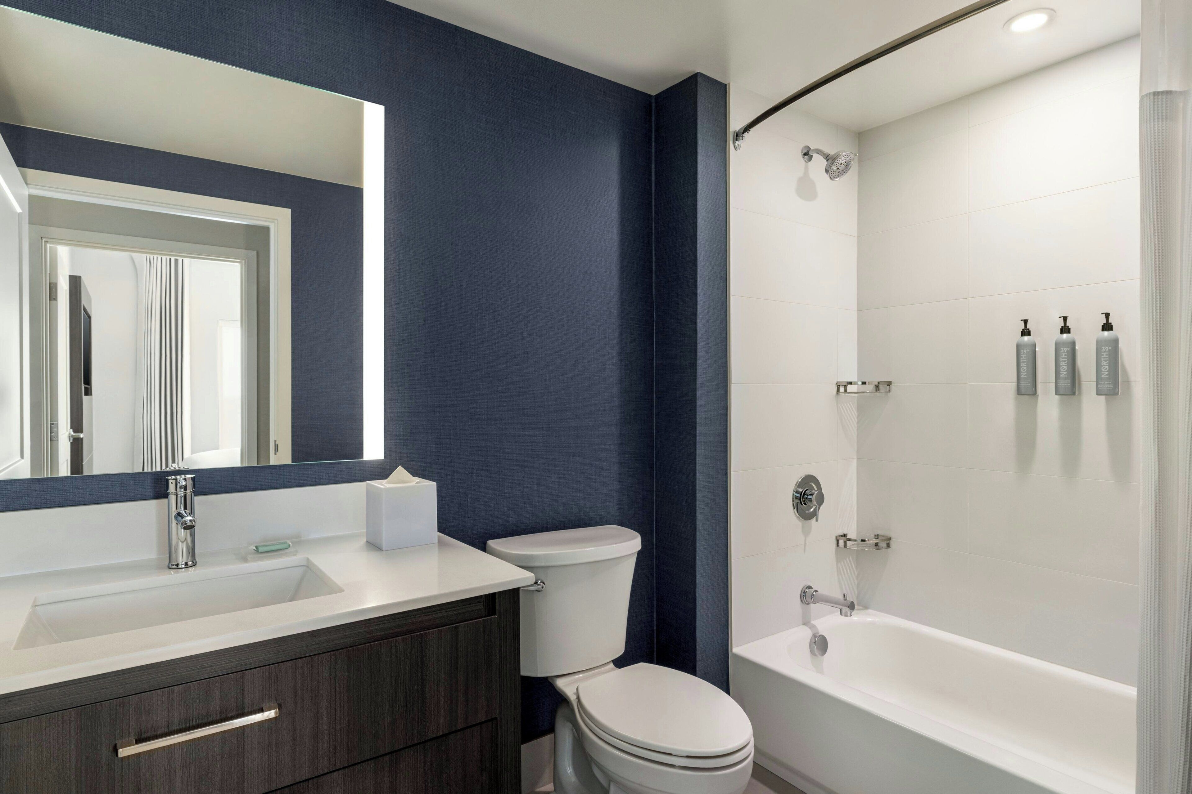 Uma casa de banho moderna com uma parede azul escura, uma sanita, uma banheira com chuveiro e uma pia branca com espelho.