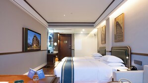 Quarto de hotel com uma cama grande, uma escrivaninha com uma chávena e um aviso, uma cadeira, uma mesa pequena com um cesto e uma parede com um quadro emoldurado.