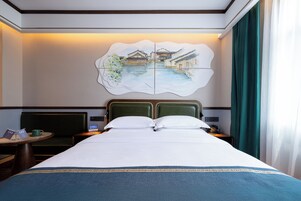 Quarto de hotel com uma cama grande, uma mesa de cabeceira, uma mesa com uma chávena e cardápio, e uma obra de arte na parede retratando um edifício tradicional.