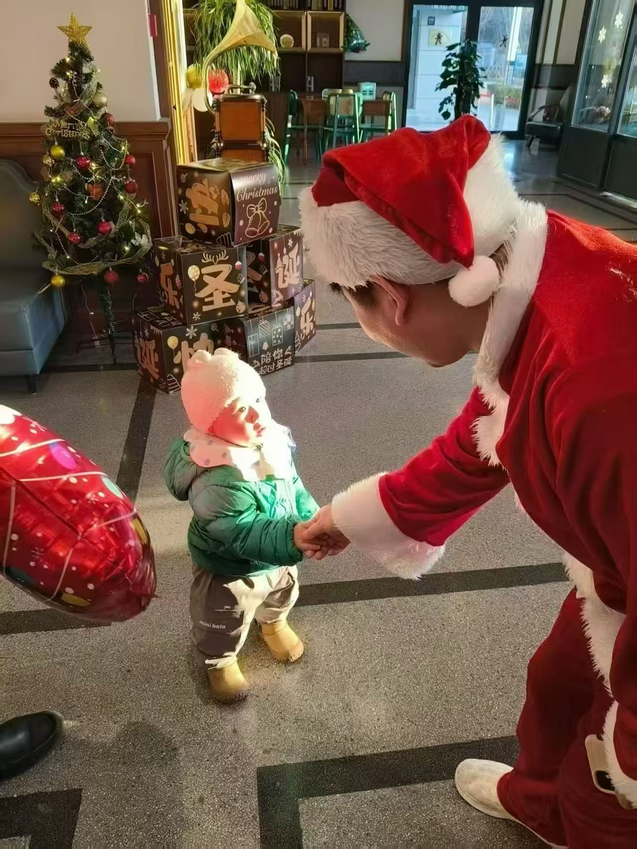 Uma pessoa vestida de Papai Noel interagindo com uma criança em um saguão decorado com árvore de Natal e caixas de presente.