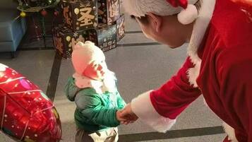Uma pessoa vestida de Papai Noel interagindo com uma criança em um saguão decorado com árvore de Natal e caixas de presente.