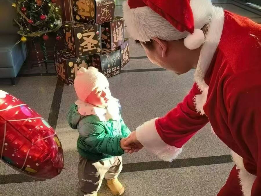 Uma pessoa vestida de Papai Noel interagindo com uma criança em um saguão decorado com árvore de Natal e caixas de presente.
