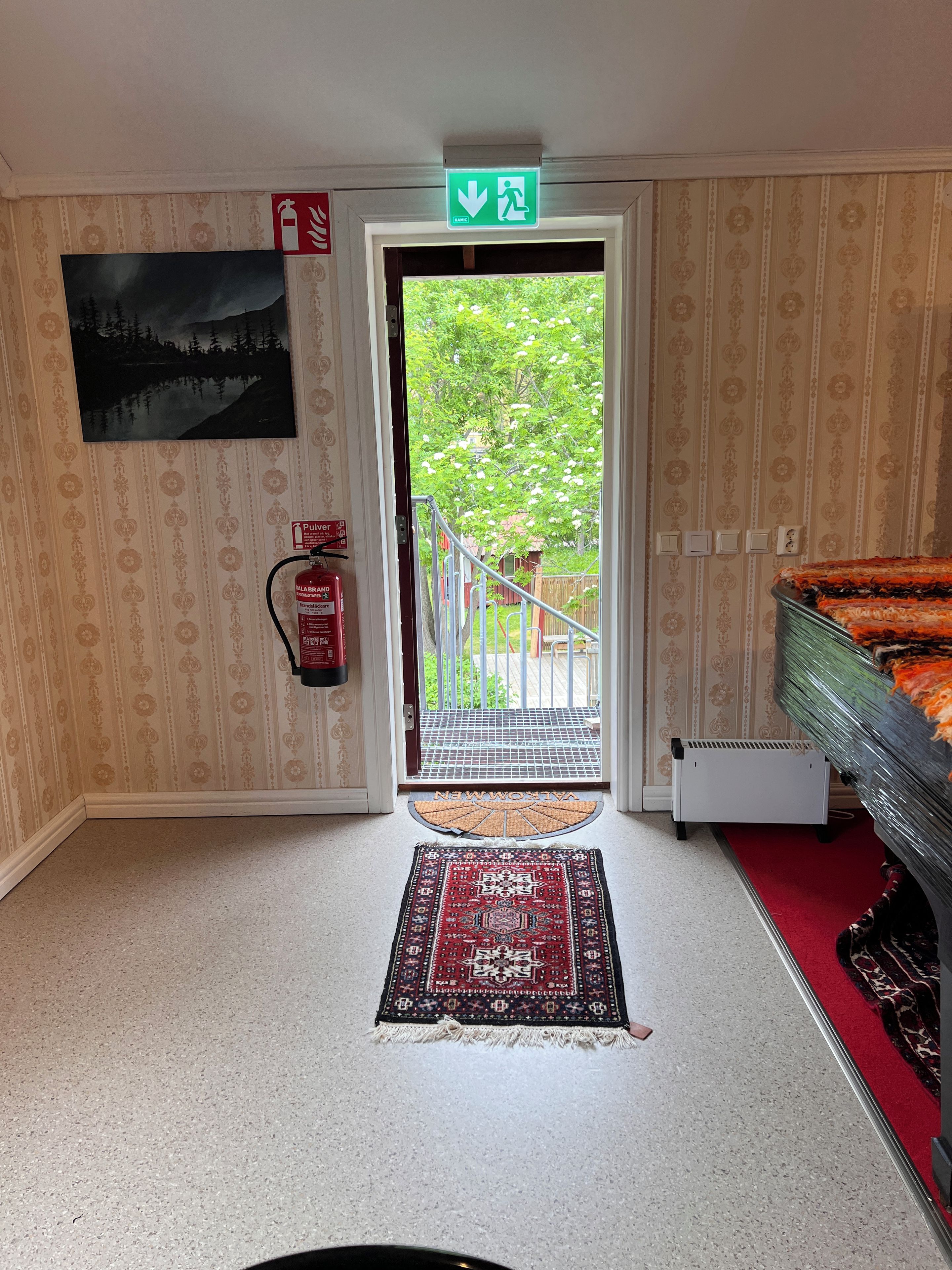 Quarto de hotel com porta para uma varanda, extintor de incêndio, quadro emoldurado e cama com cobertura vermelha.