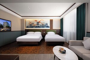 Quarto de hotel com duas camas, um sofá, uma mesa de centro e uma pintura grande de uma paisagem urbana.