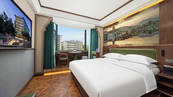 Quarto de hotel com uma cama grande, uma escrivaninha com cadeira, vista para a paisagem urbana pela janela e um quadro de uma paisagem na parede.