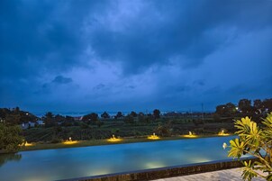 Área de piscina ao ar livre tranquila, com superfície aquática calma refletindo o céu, cercada por vegetação exuberante e vista distante de uma cidade.