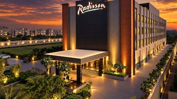 Um hotel moderno com entrada imponente, área externa ajardinada e o horizonte da cidade ao fundo.