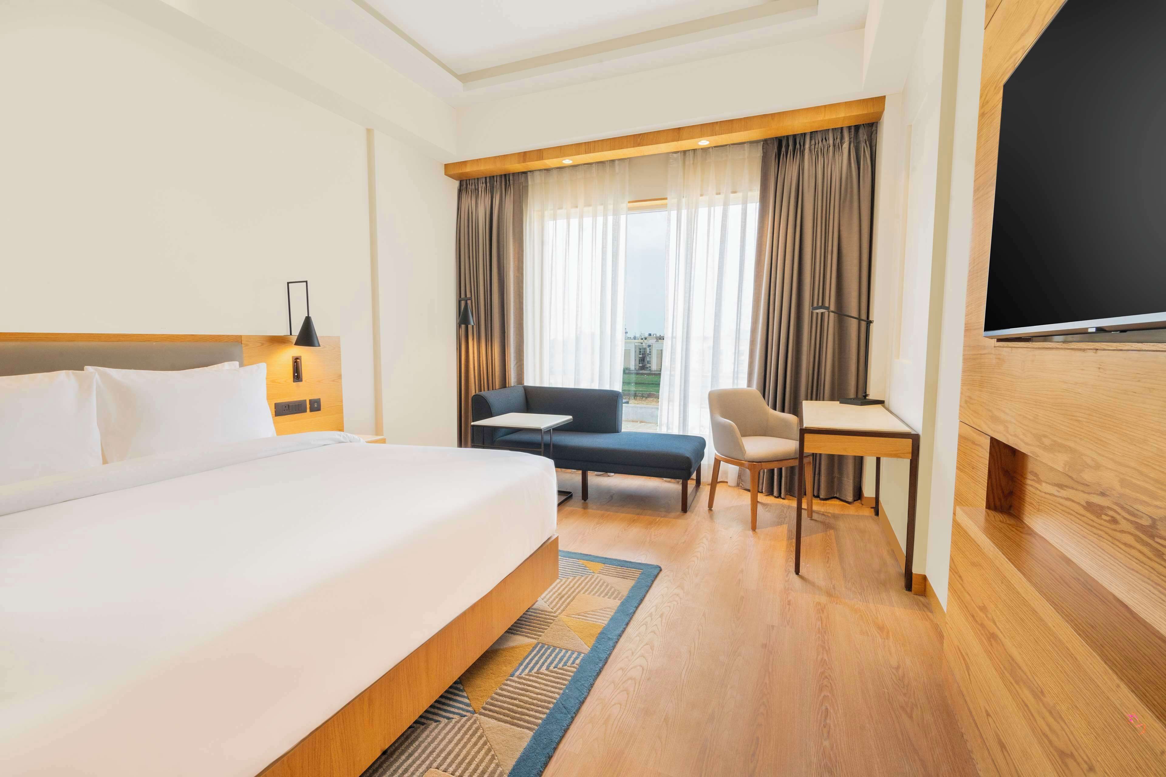 Quarto de hotel com uma cama grande, escrivaninha, cadeira, televisão e vista para o exterior.