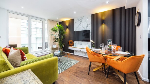 Uma sala de estar moderna com um sofá verde, cadeiras laranja e uma mesa de jantar para quatro pessoas.