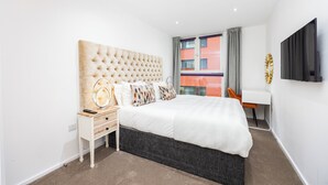 Um quarto moderno com cabeceira estofada, cama com roupa de cama branca, mesa de cabeceira com espelho e TV fixada na parede.