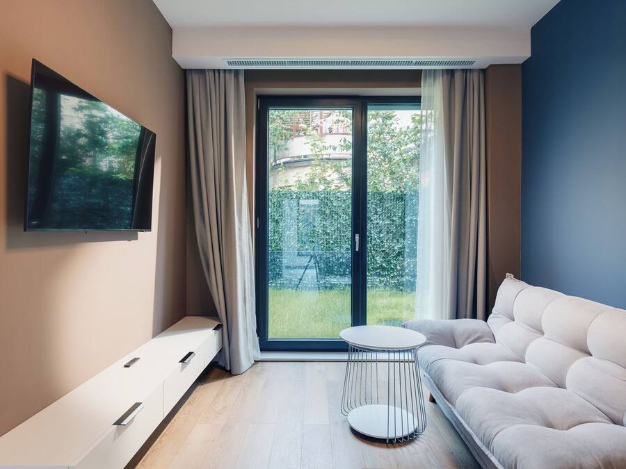 Uma sala de estar moderna com um sofá, uma televisão fixada na parede, uma porta de vidro deslizante para um jardim e uma pequena mesa redonda.
