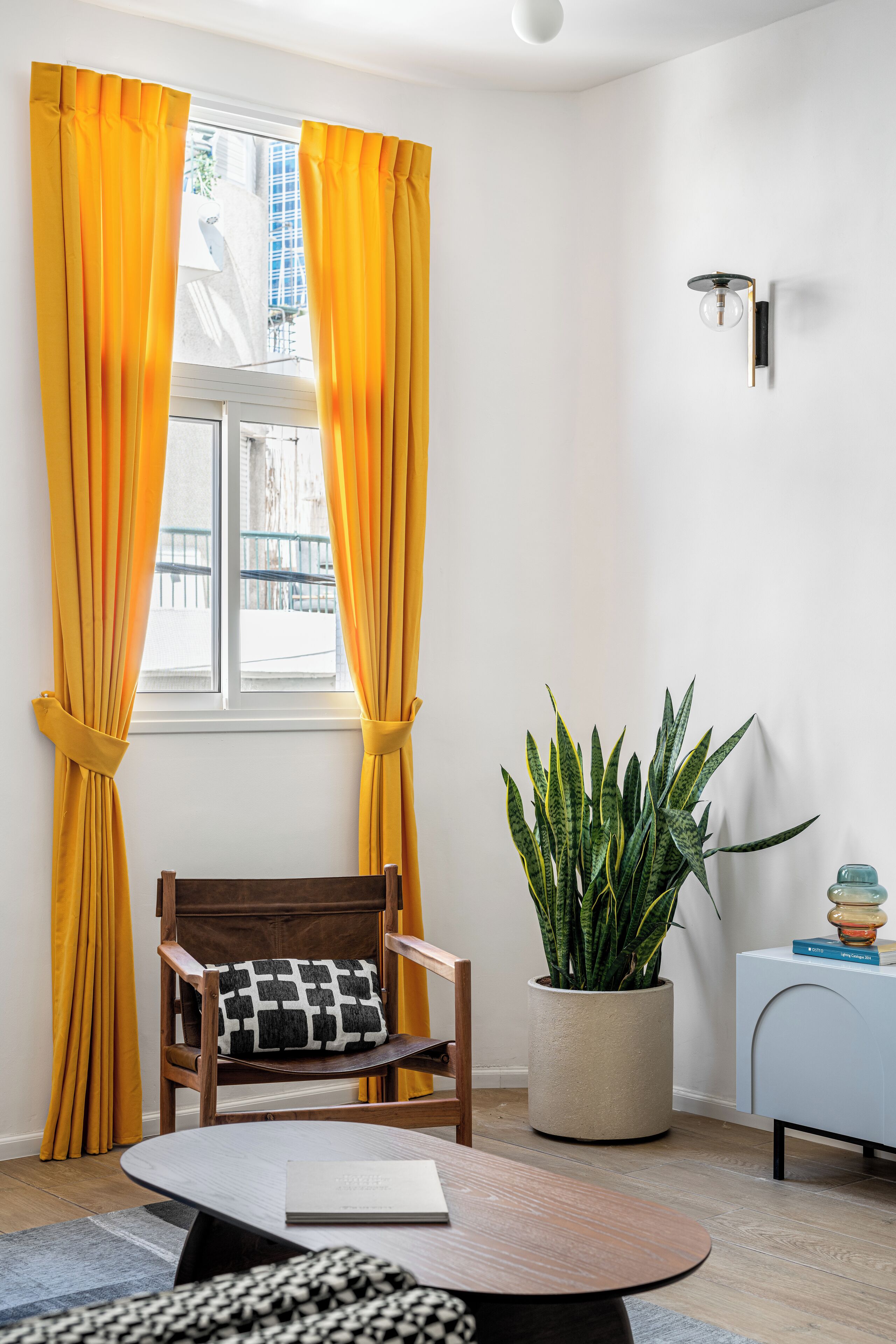 Um quarto com uma cadeira de madeira, uma mesa redonda, um vaso de planta e uma janela com cortinas amarelas.