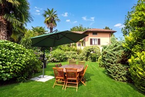 Um jardim bem cuidado com uma mesa e cadeiras para refeições, rodeado por vegetação exuberante e uma casa ao fundo.