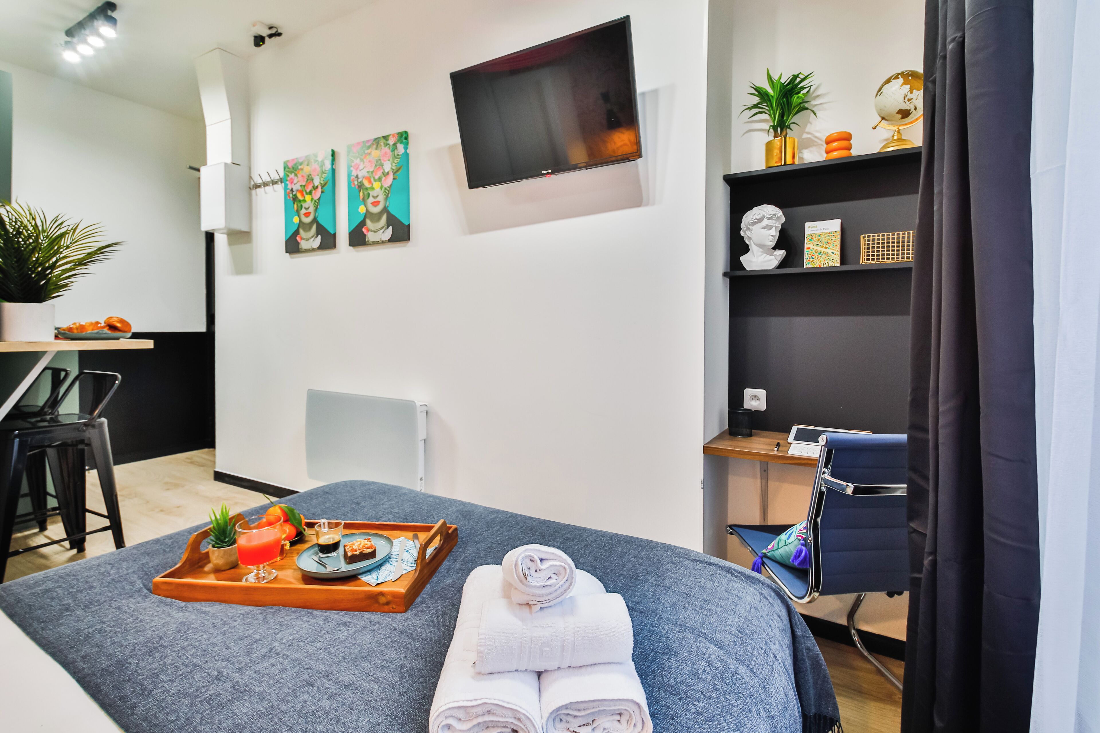 Quarto de hotel com cama, escrivaninha, cadeira, televisão e prateleira com objetos decorativos.