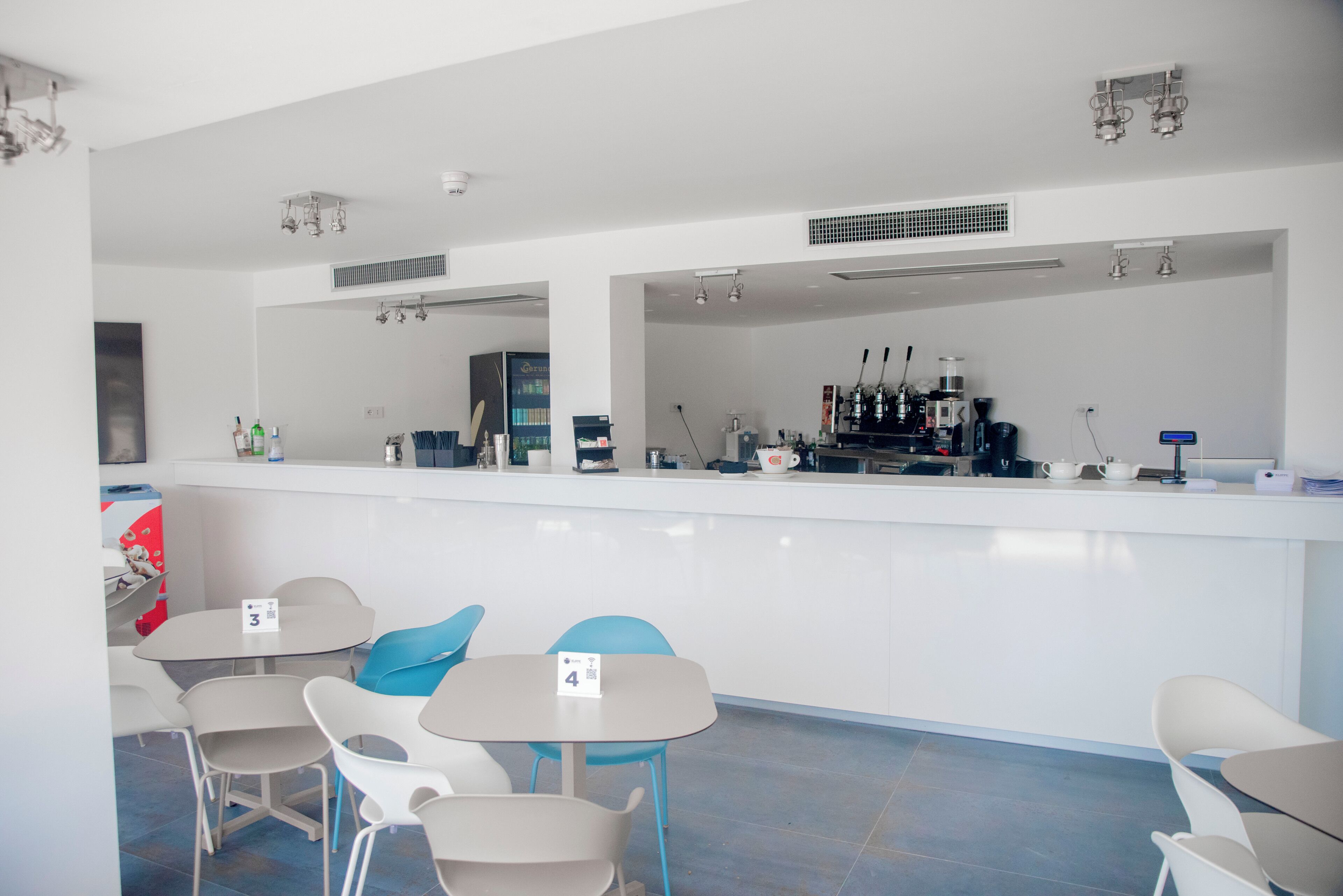 Uma área de bar limpa e moderna, com mobiliário branco e balcão. Há mesas e cadeiras, e o bar está equipado com máquinas de café e garrafas.