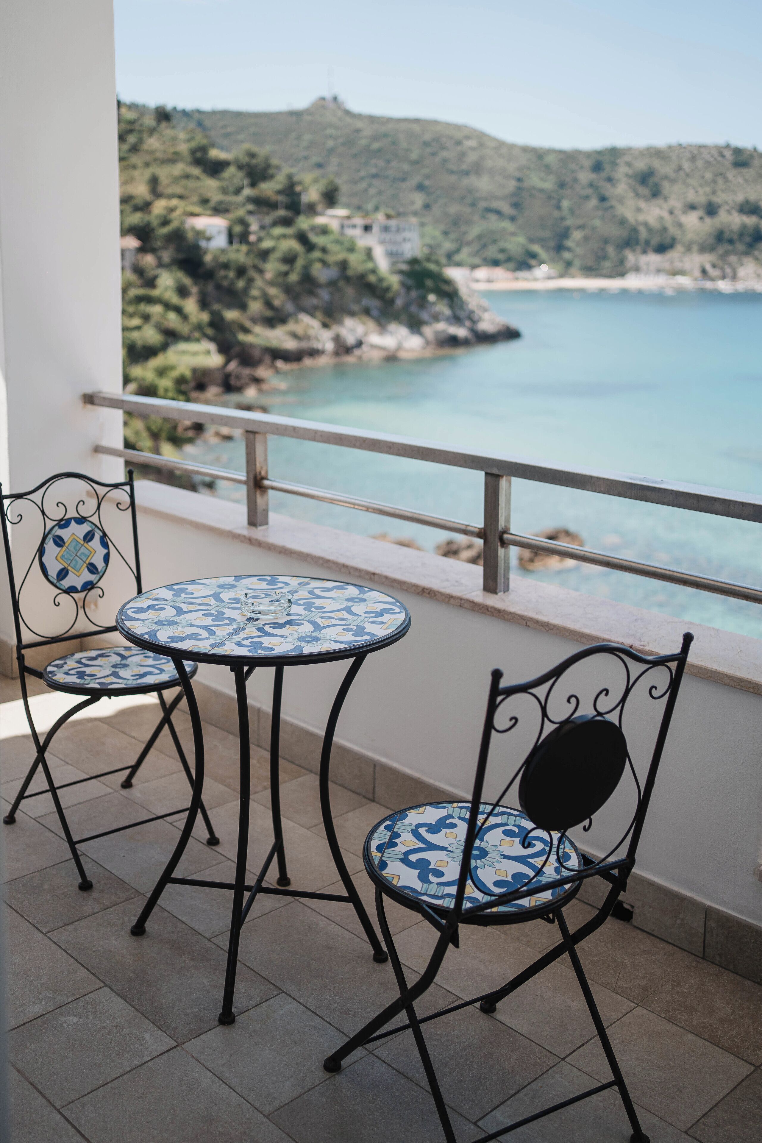 Varanda com duas cadeiras e uma mesa, com vista para a paisagem costeira com colinas e um corpo de água.