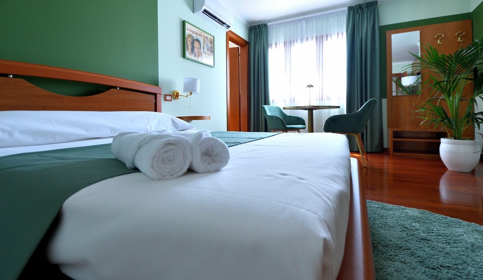 Quarto de hotel com uma cama grande, duas toalhas enroladas, uma mesa com cadeira, uma planta em vaso e um espelho.