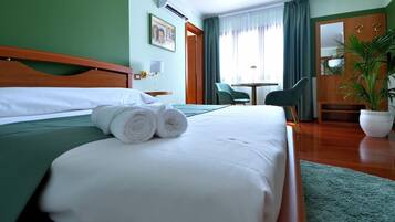 Quarto de hotel com uma cama grande, duas toalhas enroladas, uma mesa com cadeira, uma planta em vaso e um espelho.