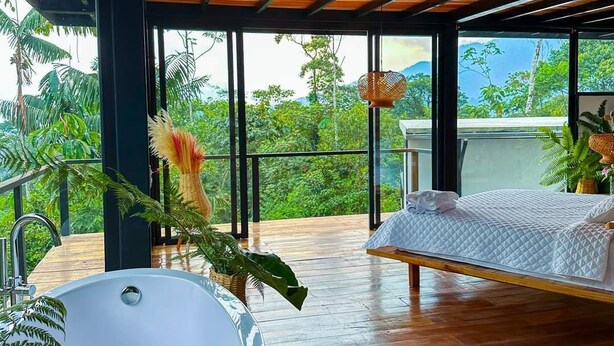 Quarto moderno com uma banheira grande, uma cama e vista para vegetação exuberante.