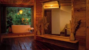 Interior de uma cabana de madeira com uma janela grande, uma banheira e um espelho refletindo o aposento.