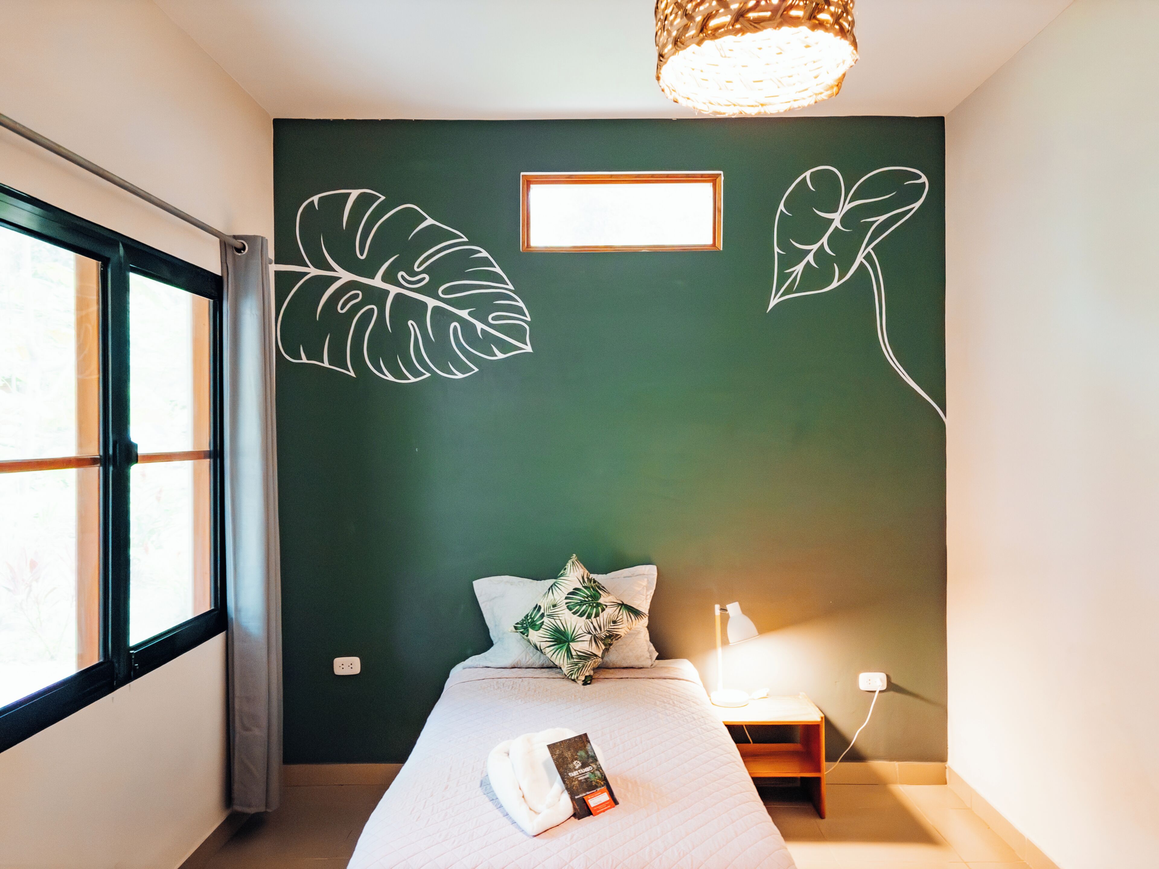 Quarto com cama, mesa de cabeceira, luminária, janela e decoração na parede.