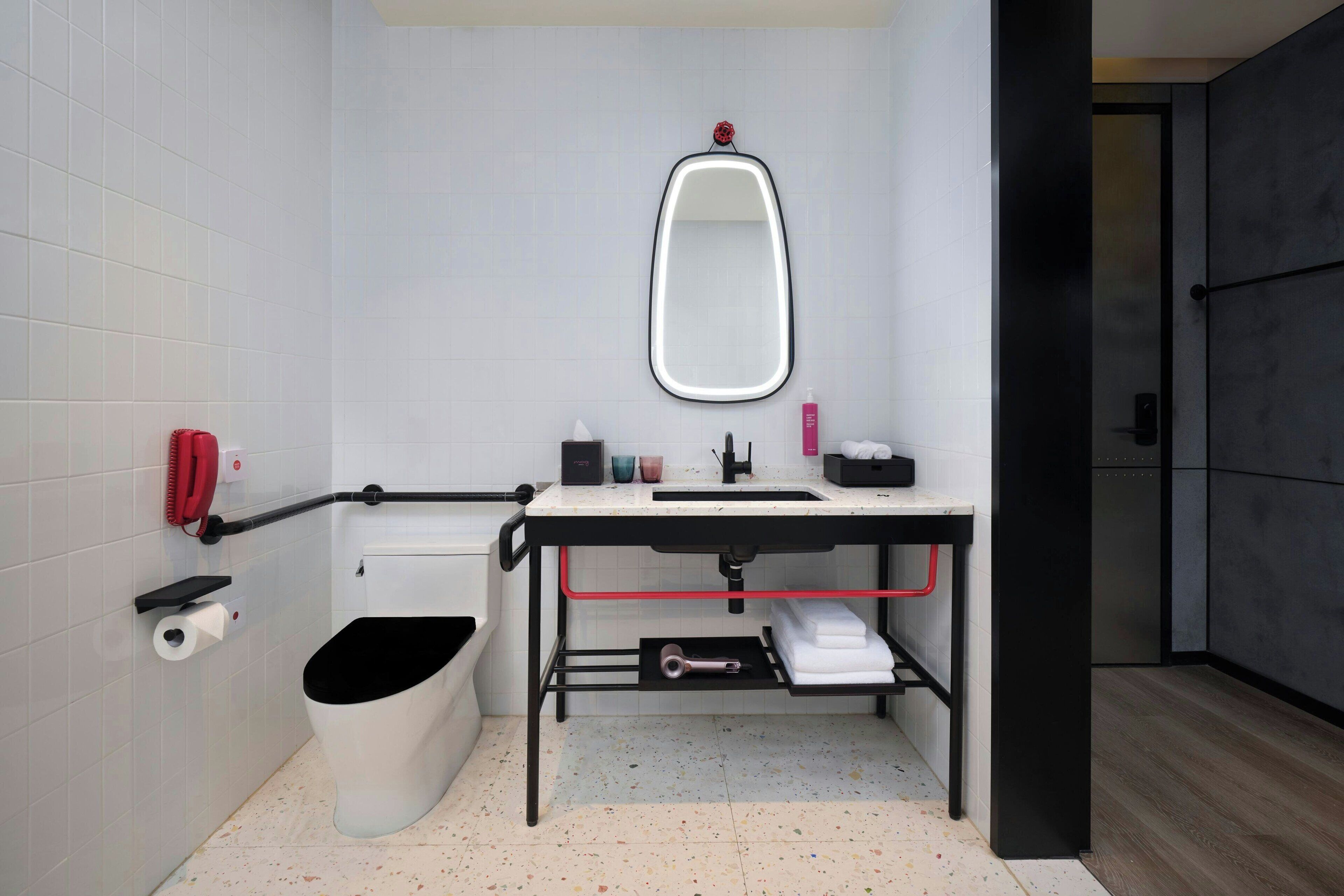 Uma casa de banho moderna com uma sanita branca, uma pia preta com espelho em cima, um porta-toalhas vermelho e um cesto de lixo preto.
