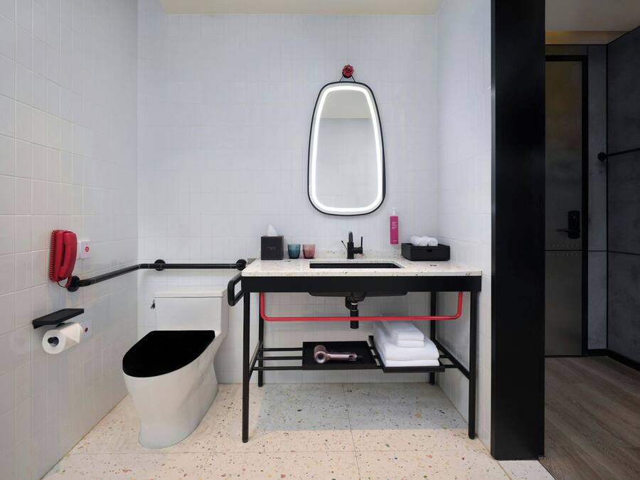 Uma casa de banho moderna com uma sanita branca, uma pia preta com espelho em cima, um porta-toalhas vermelho e um cesto de lixo preto.