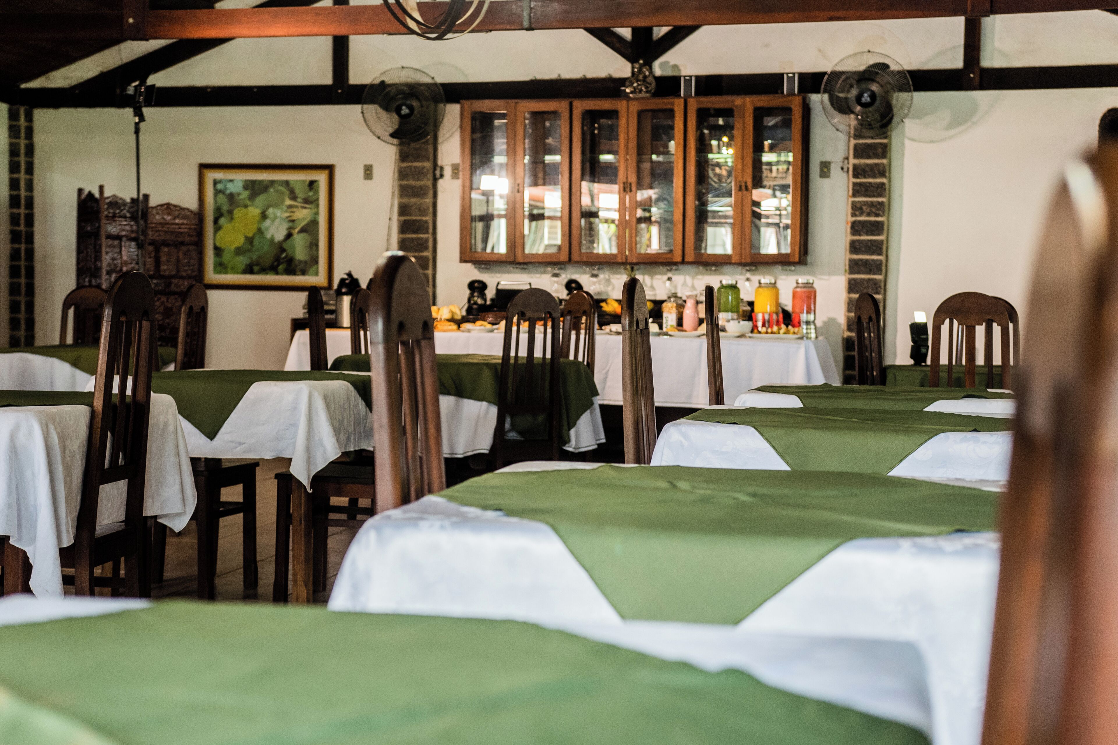 Uma sala de jantar com várias mesas cobertas por toalhas verdes e corredores de mesa brancos, cadeiras de madeira e uma parede de tijolos com uma janela e armários.