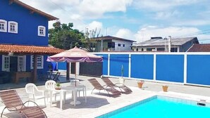 Piscina com espreguiçadeiras e uma mesa, cercada por um portão azul e edifícios.
