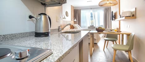 Cozinha moderna com bancada de granito, chaleira e máquina de café. Há uma área de jantar com mesa e cadeiras, e vista para a sala de estar com sofá e cortinas.