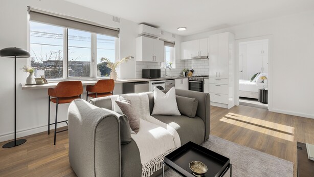 Uma sala de estar moderna com um sofá cinzento, duas poltronas laranja, uma mesa de centro e vista para a cozinha e zona de refeições através de amplas janelas.