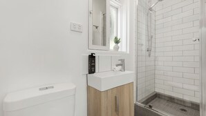 Uma casa de banho moderna com sanita branca, um armário de madeira, uma pia com espelho e uma área de duche com azulejos brancos.