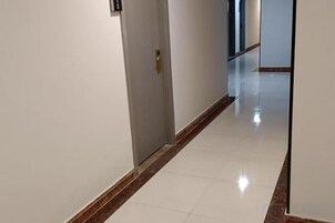 Corredor com porta à esquerda, chão revestido de azulejos refletivos e parede lisa.