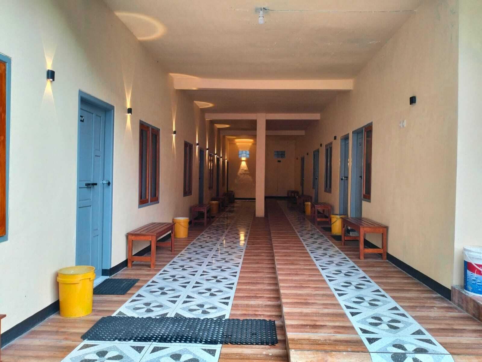 Corredor de hotel com bancos de madeira, piso de azulejos estampados e portas em ambos os lados.