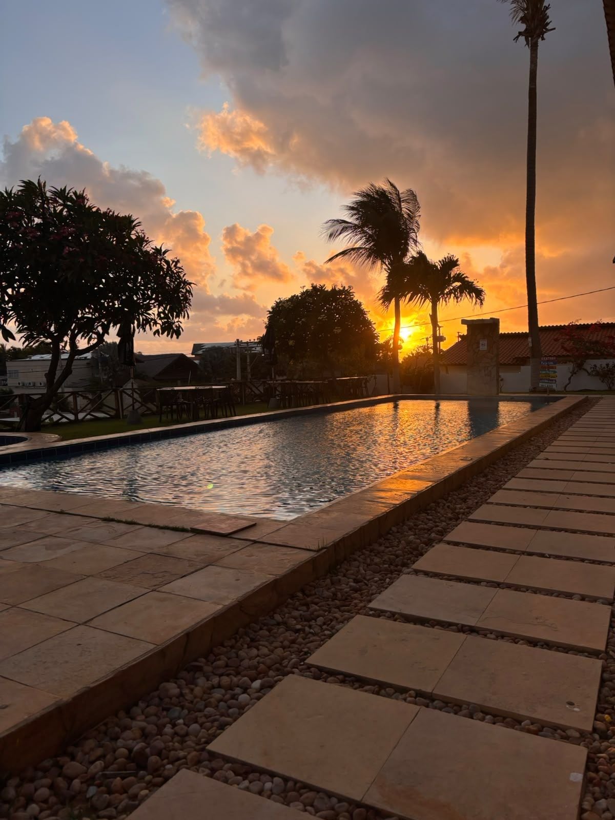 Piscina com deck revestido, rodeada por palmeiras e um pôr do sol ao fundo.