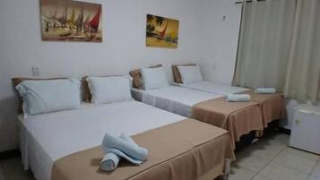 Quarto de hotel com duas camas, roupa de cama bege e branca, uma mesinha e dois quadros na parede.