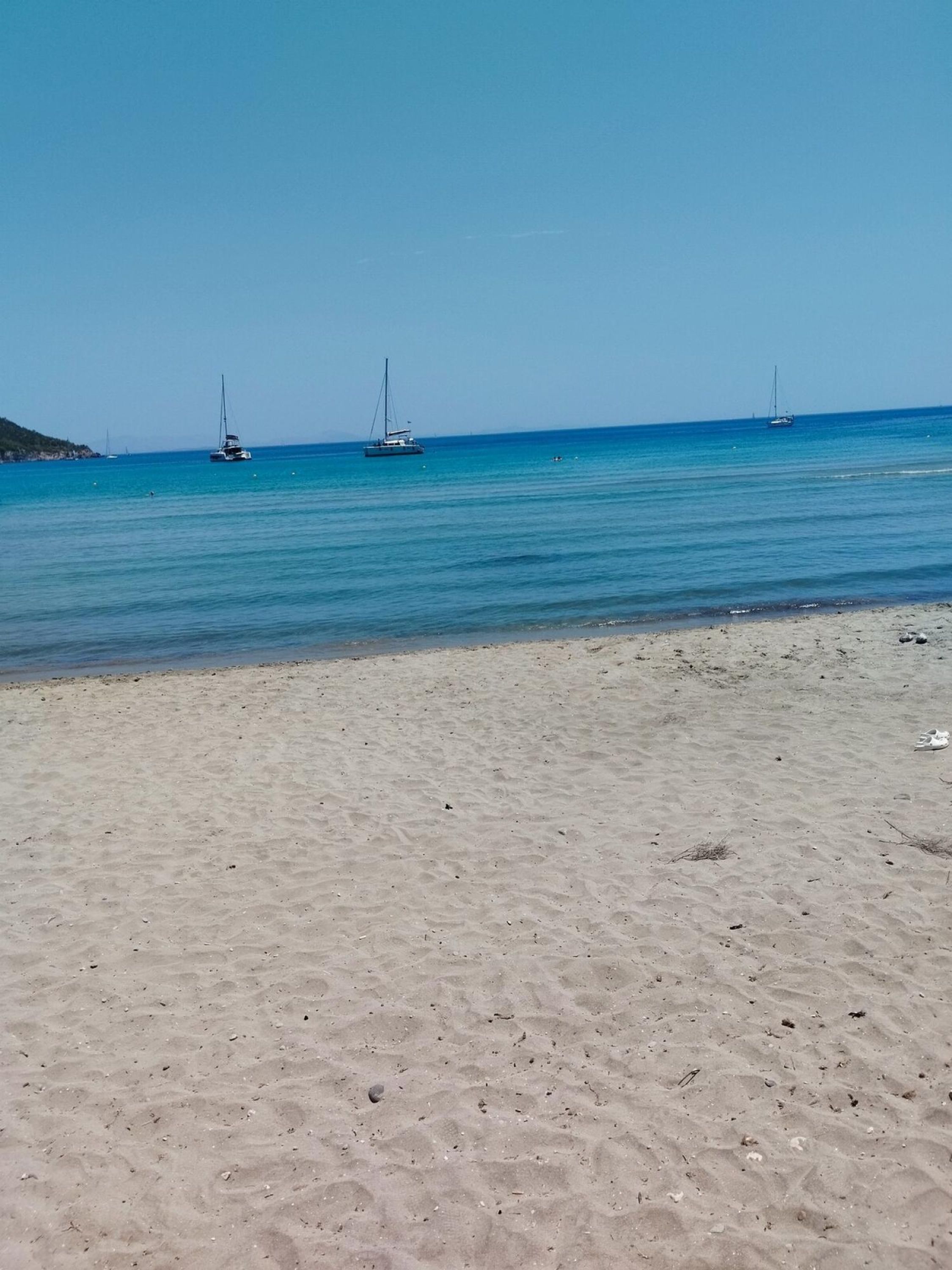 Uma praia de areia com águas calmas e veleiros ao longe.