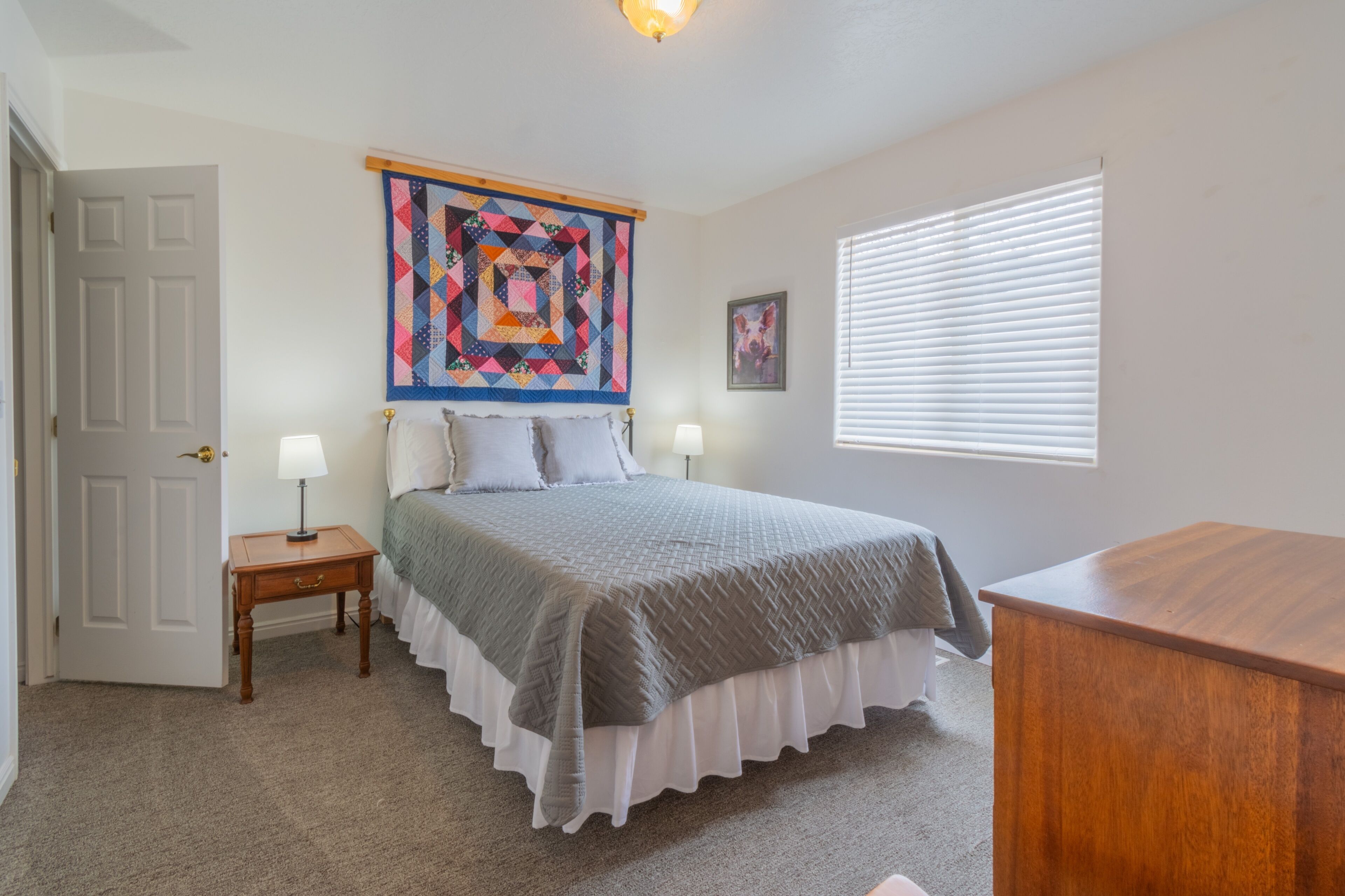 Quarto com cama, mesa de cabeceira, penteadeira, colcha e janela com persianas.